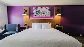 Quarto de hotel moderno com uma cama grande, paredes roxas e duas mesas de cabeceira com luminárias.