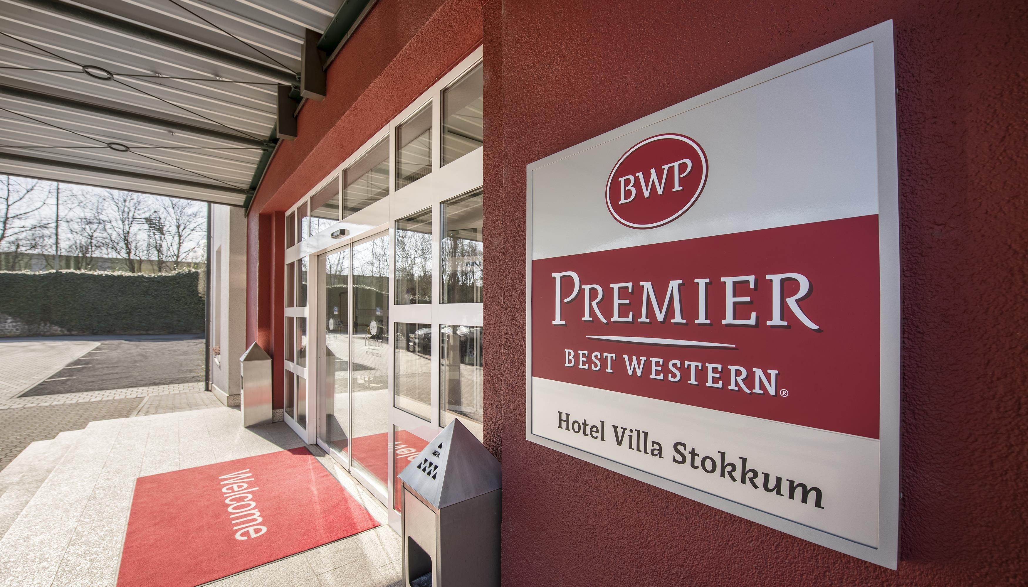 Entrada de hotel com tapete vermelho de 'Bem-vindo', placa indicando 'Premier Best Western Hotel Villa Stokkum' e área de acesso coberta.