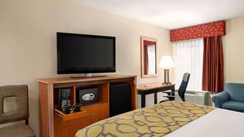 Quarto de hotel com cama, televisão, escrivaninha com luminária, cadeira e cofre.