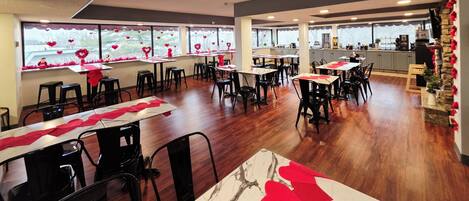 Restaurante com várias mesas e cadeiras, grandes janelas com decorações de coração e uma área de cozinha visível ao fundo.