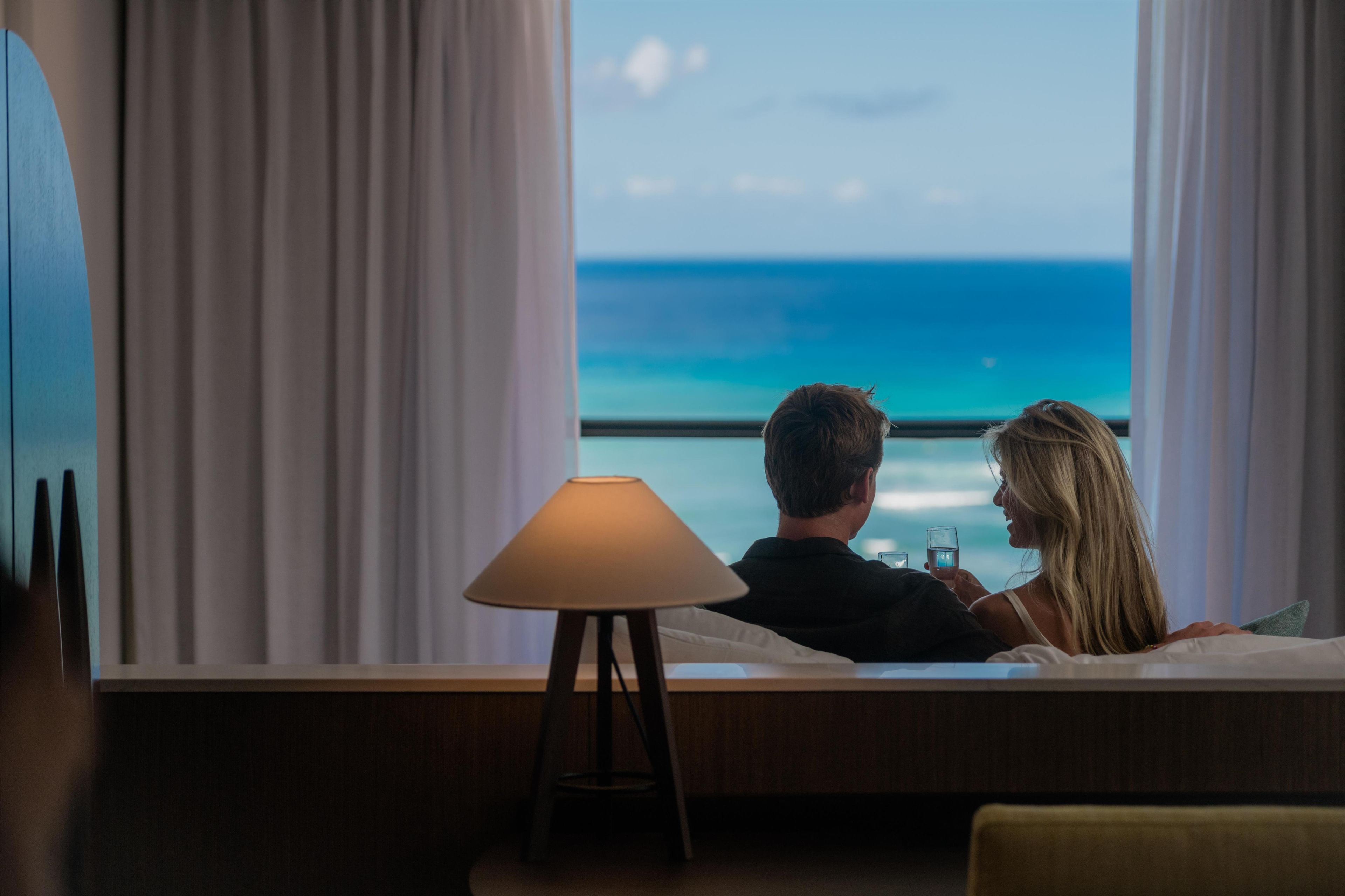 Um casal sentado perto de uma janela com vista para o oceano, um abajur na mesa e uma cama com cabeceira branca.