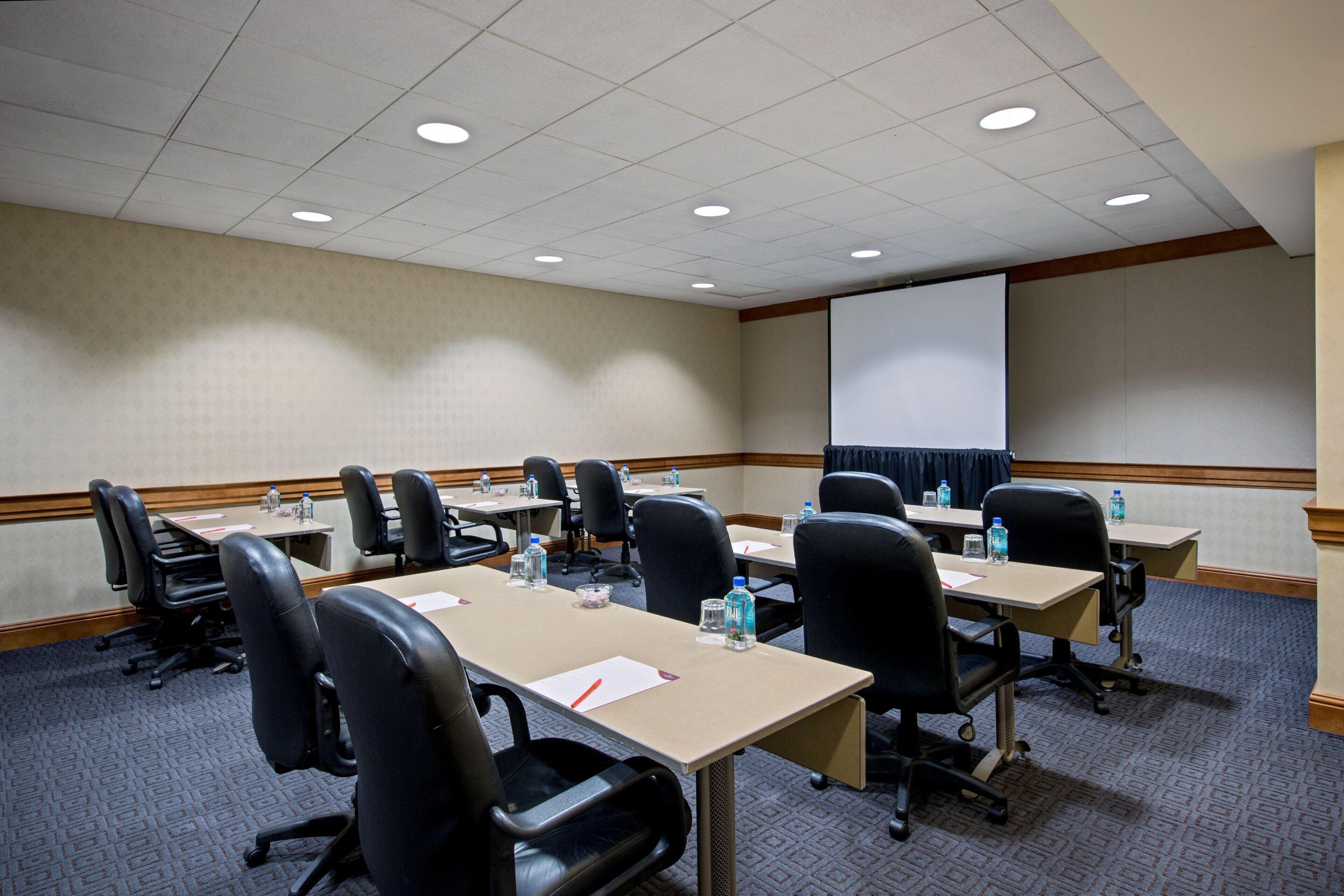 Sala de conferências com várias mesas e cadeiras, equipada com garrafas de água e papéis, e uma tela de projeção na parede.