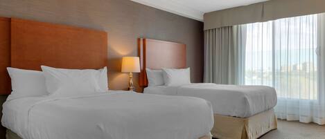 Quarto de hotel com duas camas, uma mesa de cabeceira com abajur e uma janela grande com cortinas.