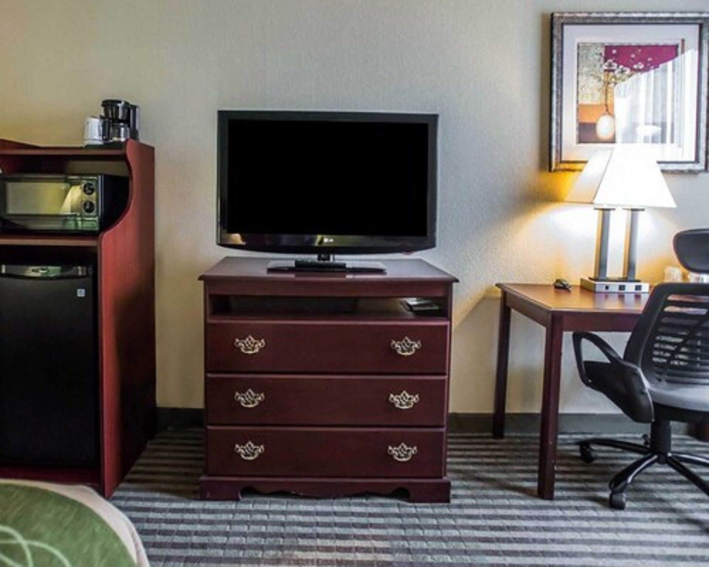 Quarto de hotel com televisão de tela plana em cômoda de madeira, escrivaninha com luminária e uma cadeira.
