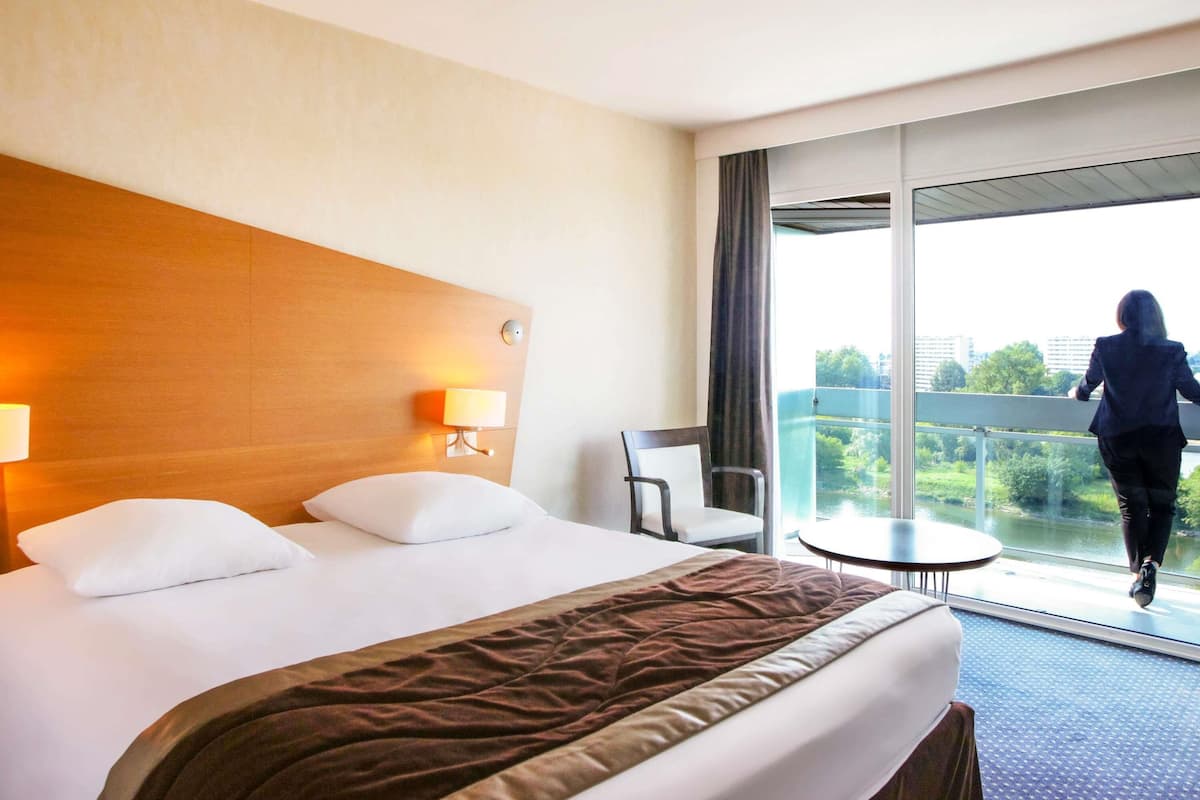 Quarto de hotel com cama, luminária de cabeceira, cadeira, mesa pequena e vista para uma varanda.