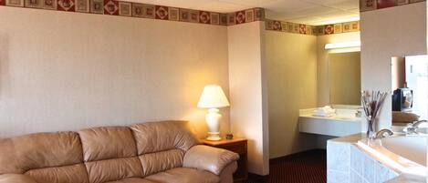 Quarto de hotel com sofá de couro, luminária de cabeceira, banheiro com pia e espelho, e piso ladrilhado.