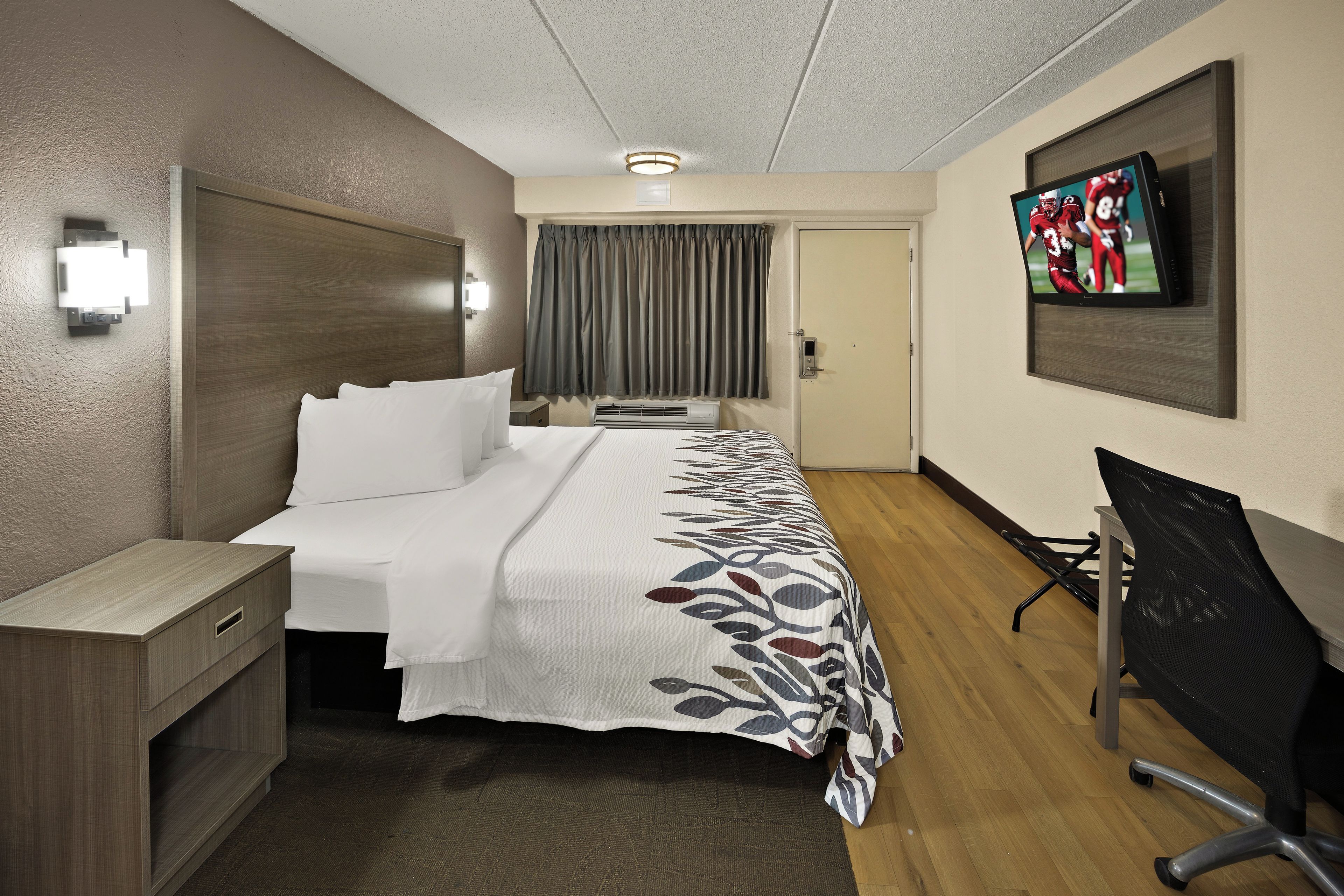 Quarto de hotel com cama, mesa de cabeceira, escrivaninha, cadeira, televisão e porta.