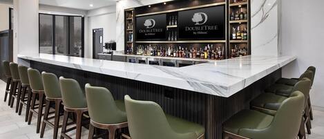 Uma área de bar com bancada de mármore, diversas garrafas de bebidas e banquetas estofadas verdes.