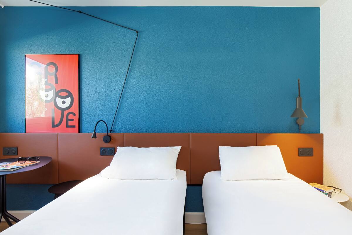 Quarto de hotel com duas camas, uma mesa pequena, uma luminária e uma obra de arte na parede.