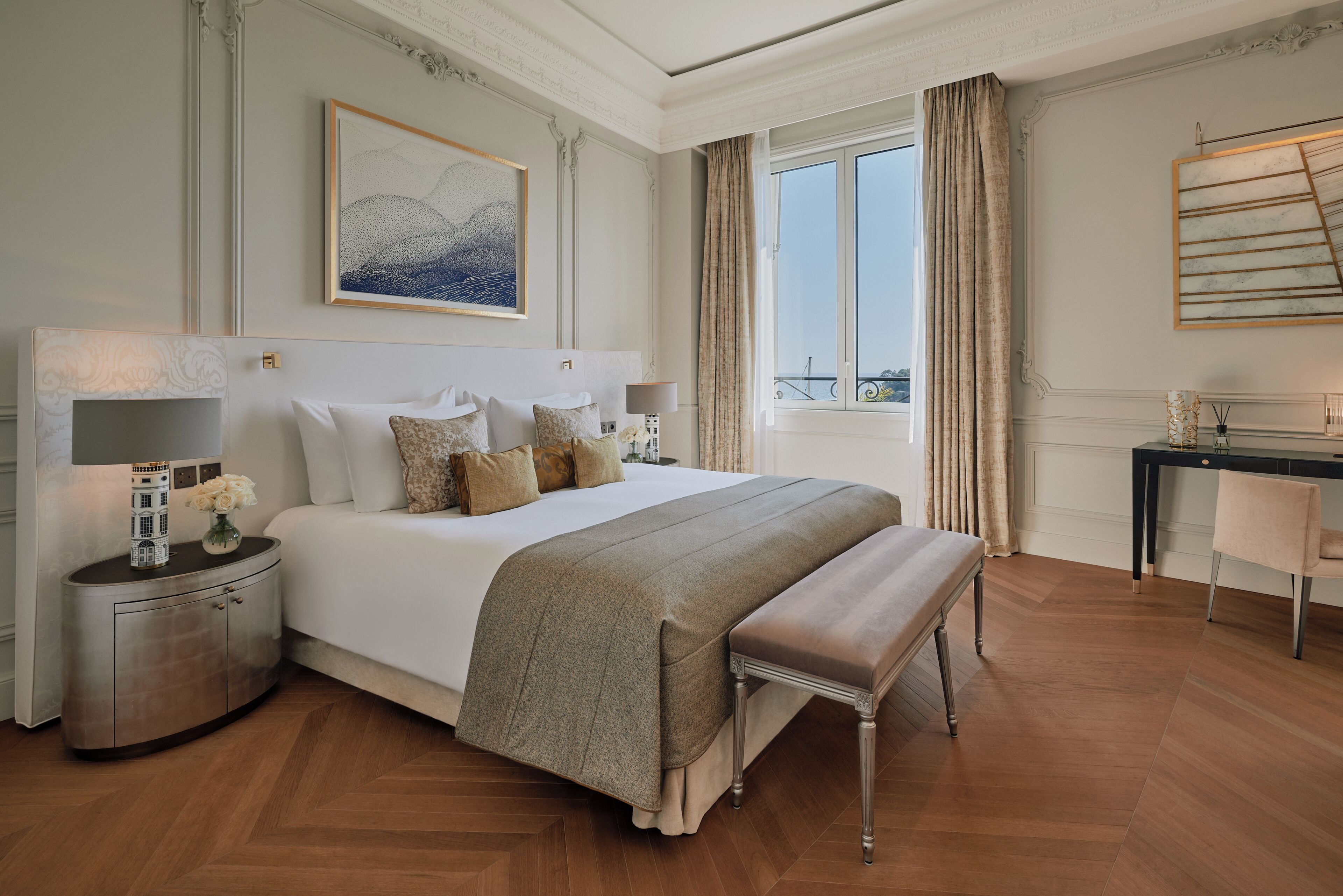 Quarto com uma cama grande, mesas de cabeceira, um banco, uma escrivaninha e vista para o exterior.