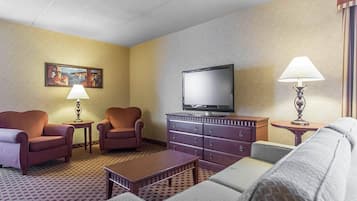 Quarto de hotel com sofá, duas poltronas, televisão, luminária e um quadro na parede.