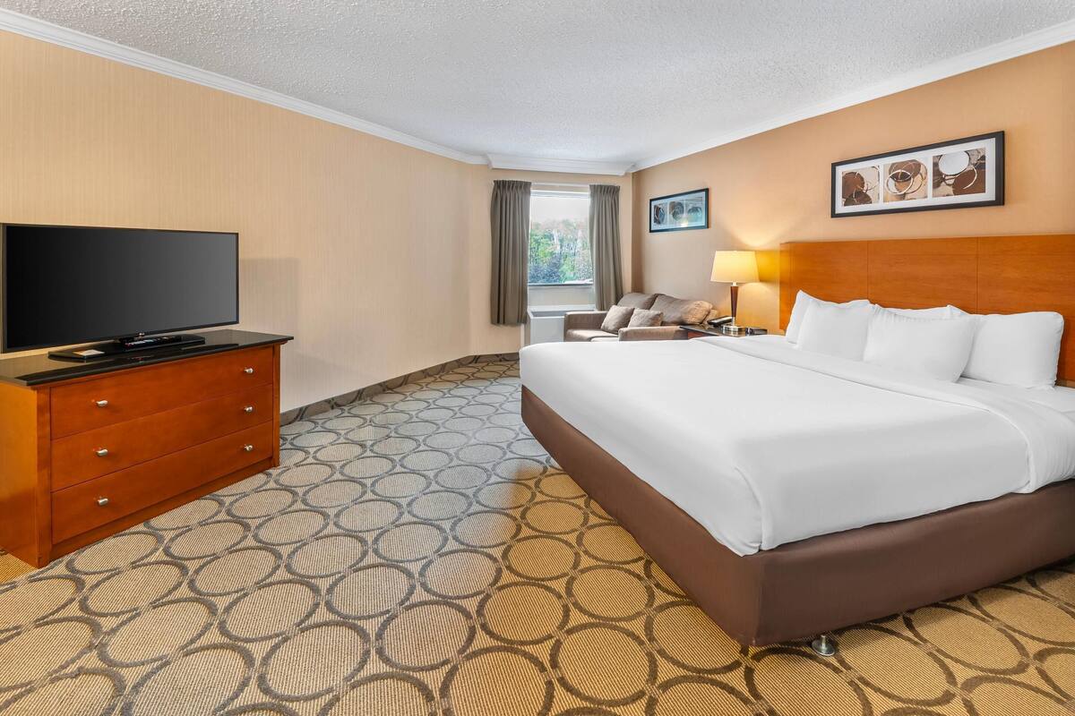 Quarto de hotel com uma cama grande, televisão de tela plana, cómoda de madeira e janela com cortinas.