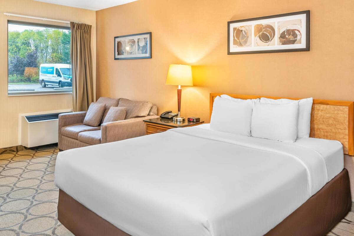 Quarto de hotel com uma cama grande, uma cadeira, uma escrivaninha com luminária e uma janela com vista para uma van lá fora.