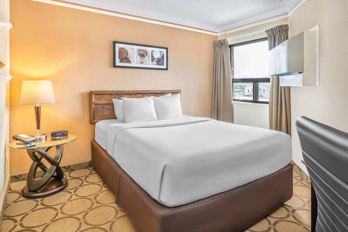 Quarto de hotel com uma cama, uma mesa de cabeceira com abajur, um quadro na parede e uma janela com cortinas.