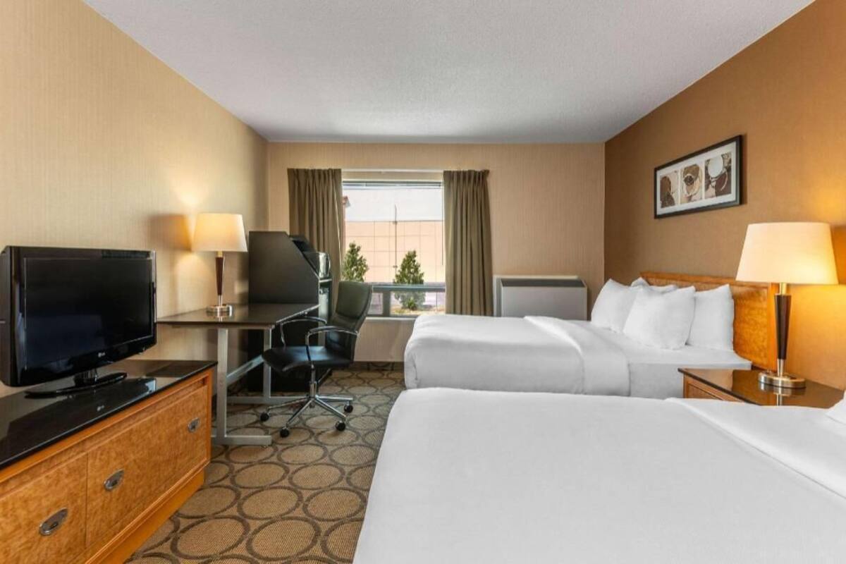 Quarto de hotel com cama, escrivaninha com televisão, cadeira, cômoda e janela com cortinas.