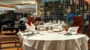 Um restaurante com piano, mesas preparadas com copos de vinho e talheres, e uma parede repleta de garrafas de vinho.