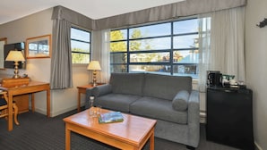 Quarto de hotel com sofá cinzento, mesa de centro de madeira, escrivaninha com luminária, frigorífico e janelas amplas com cortinas.