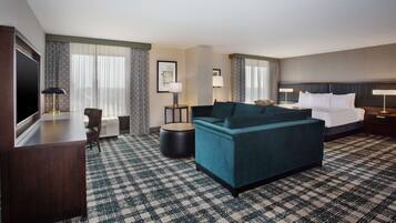 Quarto de hotel com carpete xadrez, sofá, cama, escrivaninha com televisão e abajur.