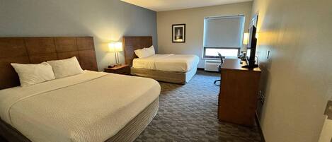 Quarto de hotel com duas camas, uma escrivaninha, uma cadeira, um abajur, um quadro na parede e uma janela com persianas.