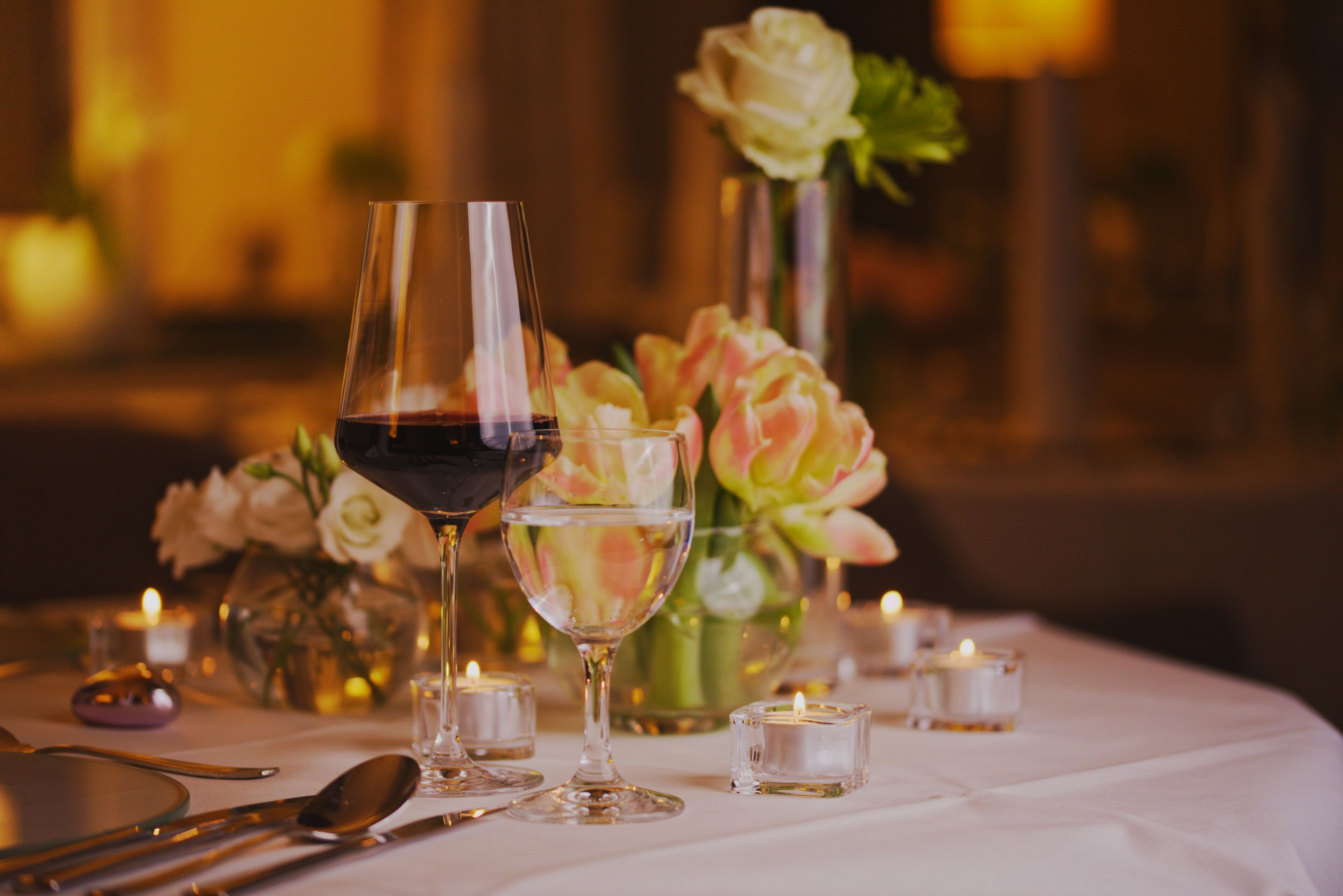 Uma mesa posta com taças de vinho, talheres e velas, rodeada por flores e vasos.