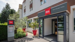 Entrada de um hotel moderno com porta de vidro, placa vermelha com o nome 'ibis' e uma pequena área externa com plantas em vasos.