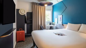 Quarto de hotel com cama, escrivaninha, um laptop, mala, televisão e janela com cortinas.