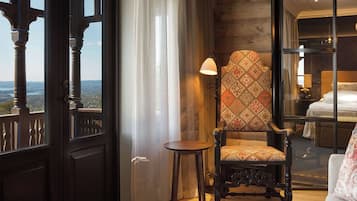 Quarto com parede revestida de madeira, uma janela ampla com vista, uma cadeira de madeira, uma mesa redonda pequena e uma cama com colcha branca.