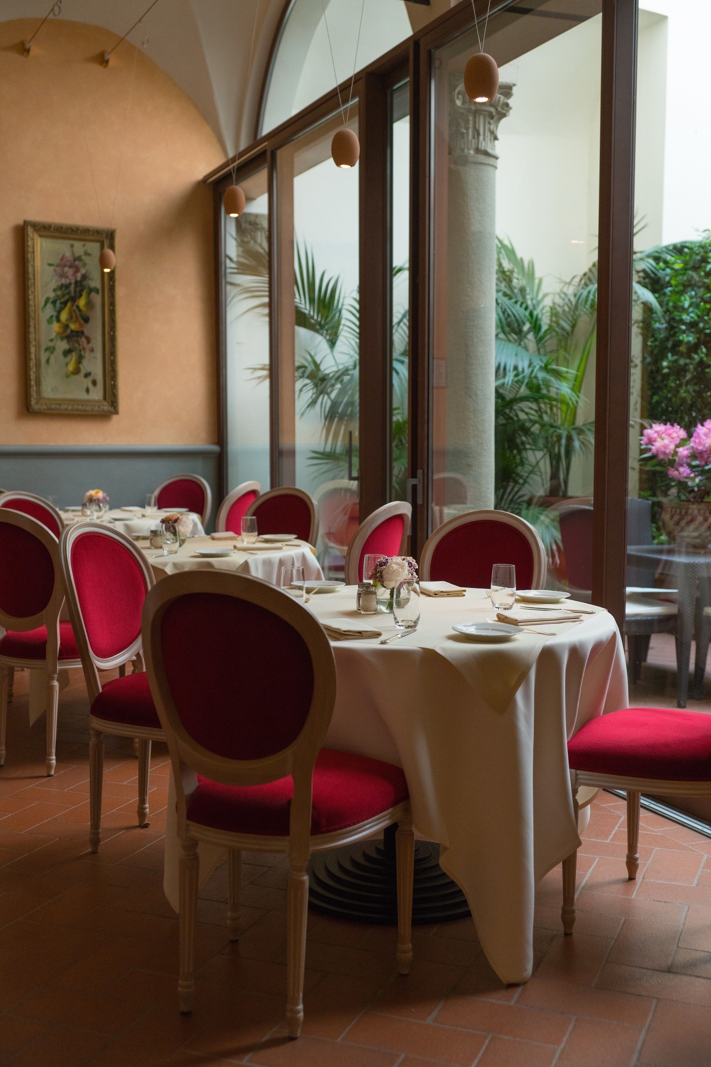 Uma sala de jantar com cadeiras estofadas vermelhas, toalhas de mesa brancas e vista para o verde através de grandes janelas.