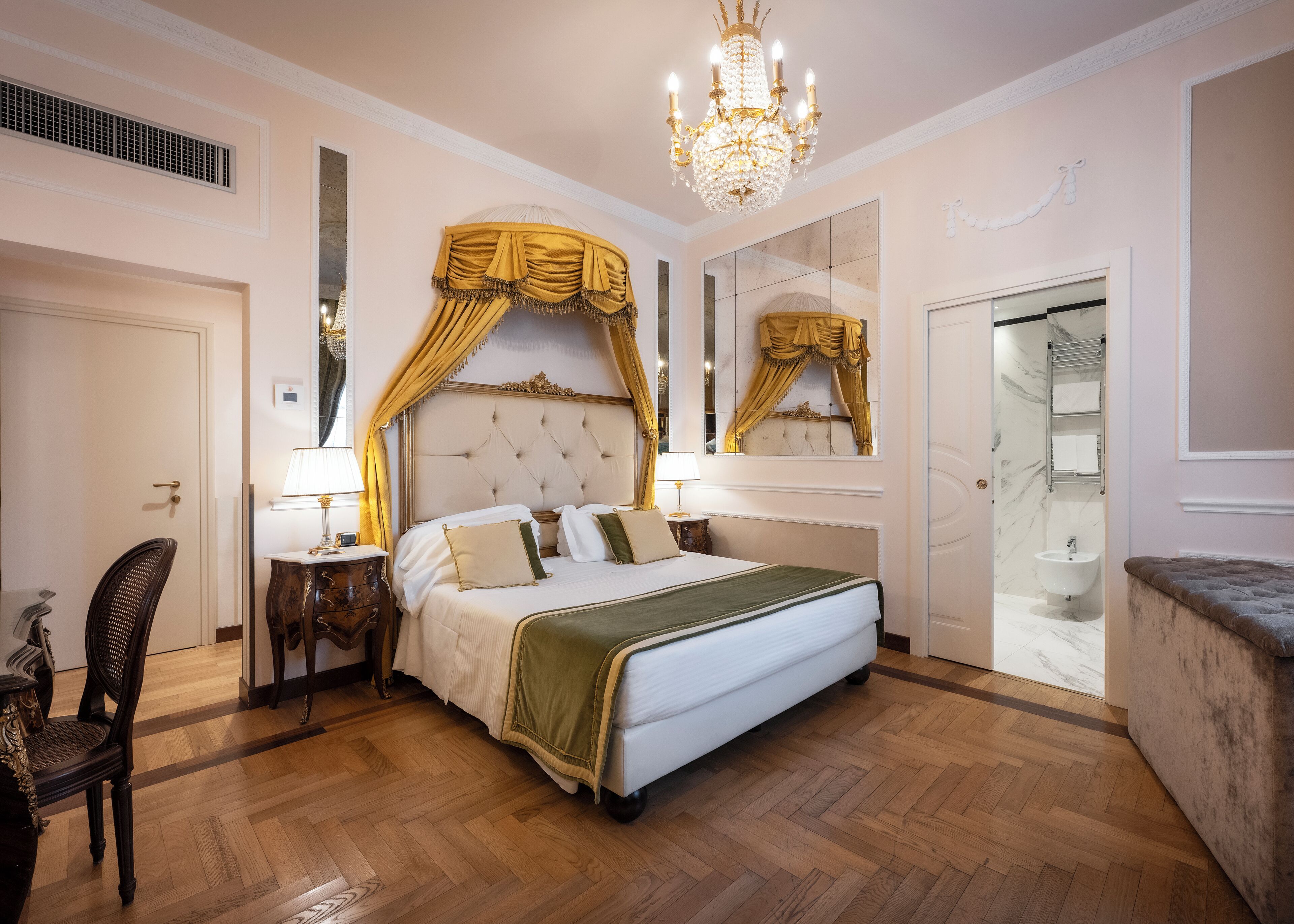 Quarto com cama grande, um candelabro, um espelho e um banheiro visível através de uma porta aberta.