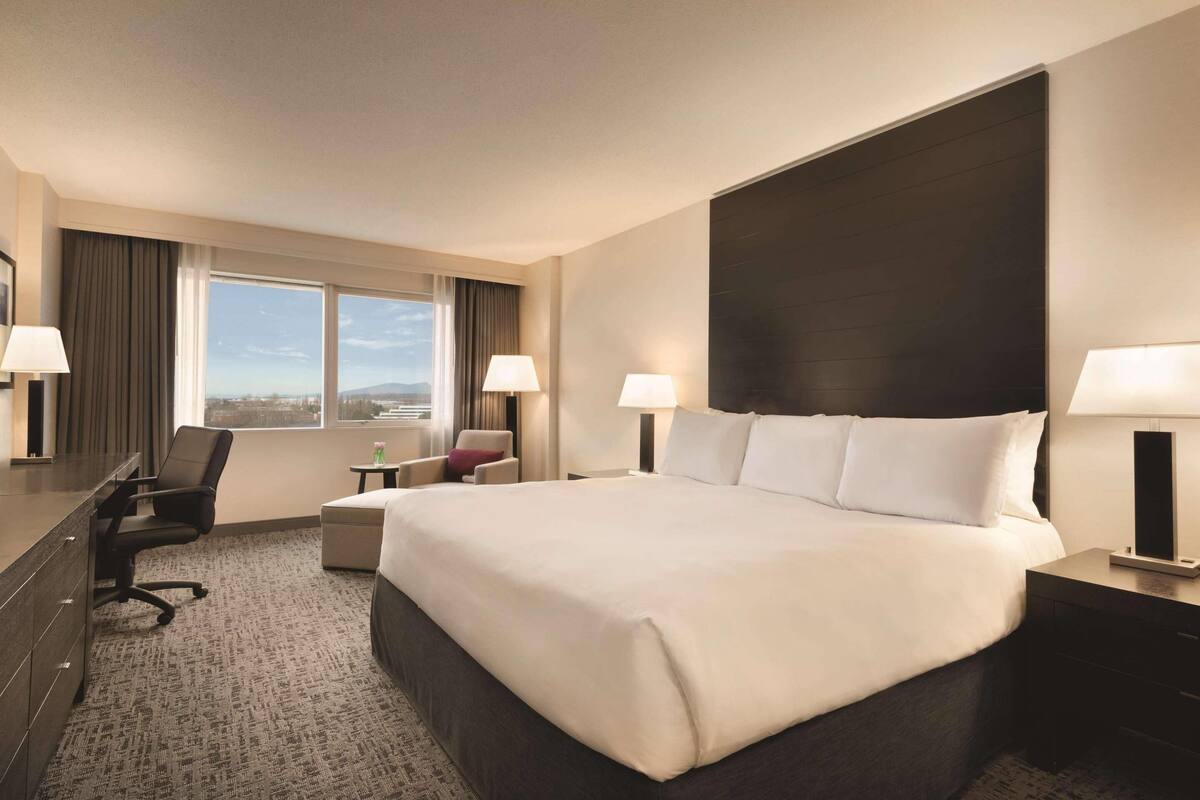 Quarto de hotel com uma cama grande, duas mesas de cabeceira com luminárias, uma escrivaninha com cadeira, uma mesinha e vista para a cidade.