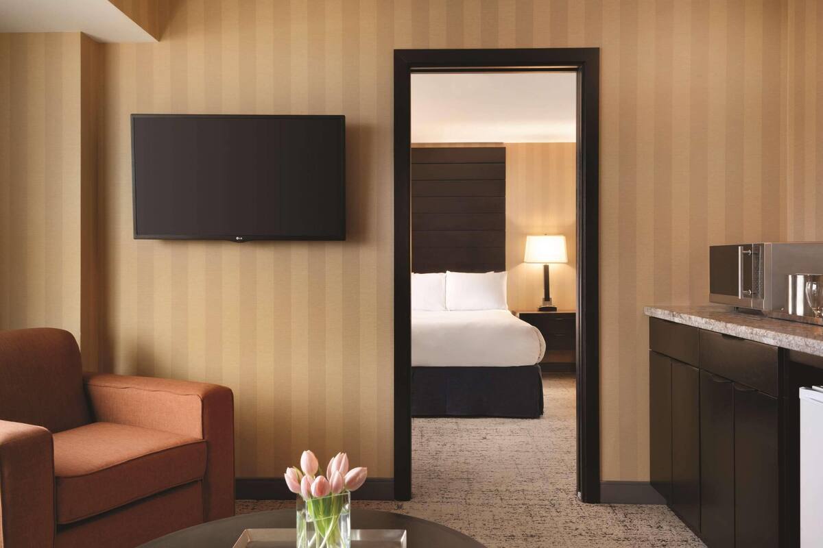 Quarto de hotel com televisão de tela plana, uma cama, mesa de cabeceira, abajur, cafeteira e área de refeições com micro-ondas.