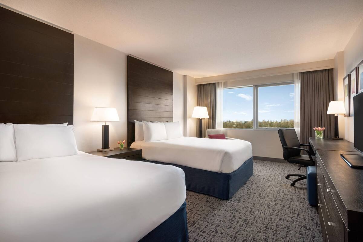 Quarto de hotel com duas camas, uma escrivaninha, uma cadeira e uma janela grande.