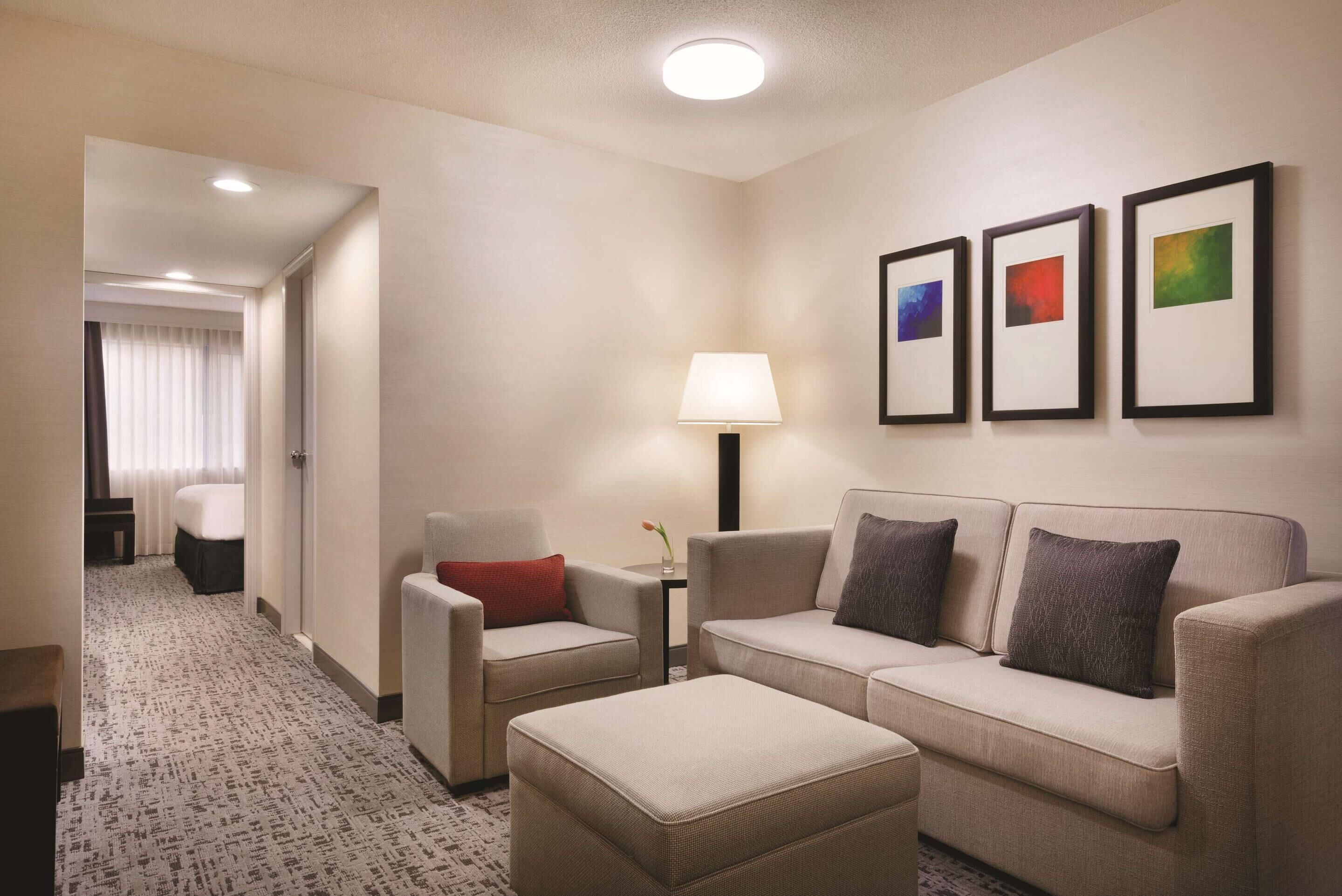 Quarto de hotel com sofá, poltrona, banquinho e abajur. Há quadros nas paredes.