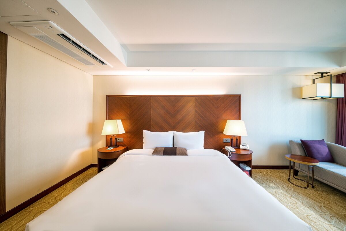 Quarto de hotel com uma cama grande, duas mesas de cabeceira com abajures, um sofá e um painel de cabeceira de madeira.