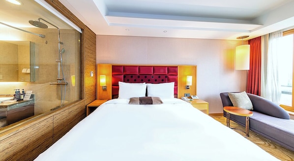 Quarto de hotel com uma cama grande, cabeceira estofada vermelha, um sofá, uma mesinha e box de vidro.