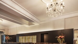 Uma área de recepção com piso de mármore, balcão escuro, lustre e uma parede com vitrine iluminada.