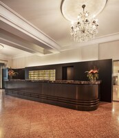Uma área de recepção com piso de mármore, balcão escuro, lustre e uma parede com vitrine iluminada.