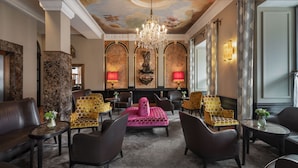 Um lobby luxuoso com sofás de couro, cadeiras estampadas, um banquinho rosa central e um candelabro.