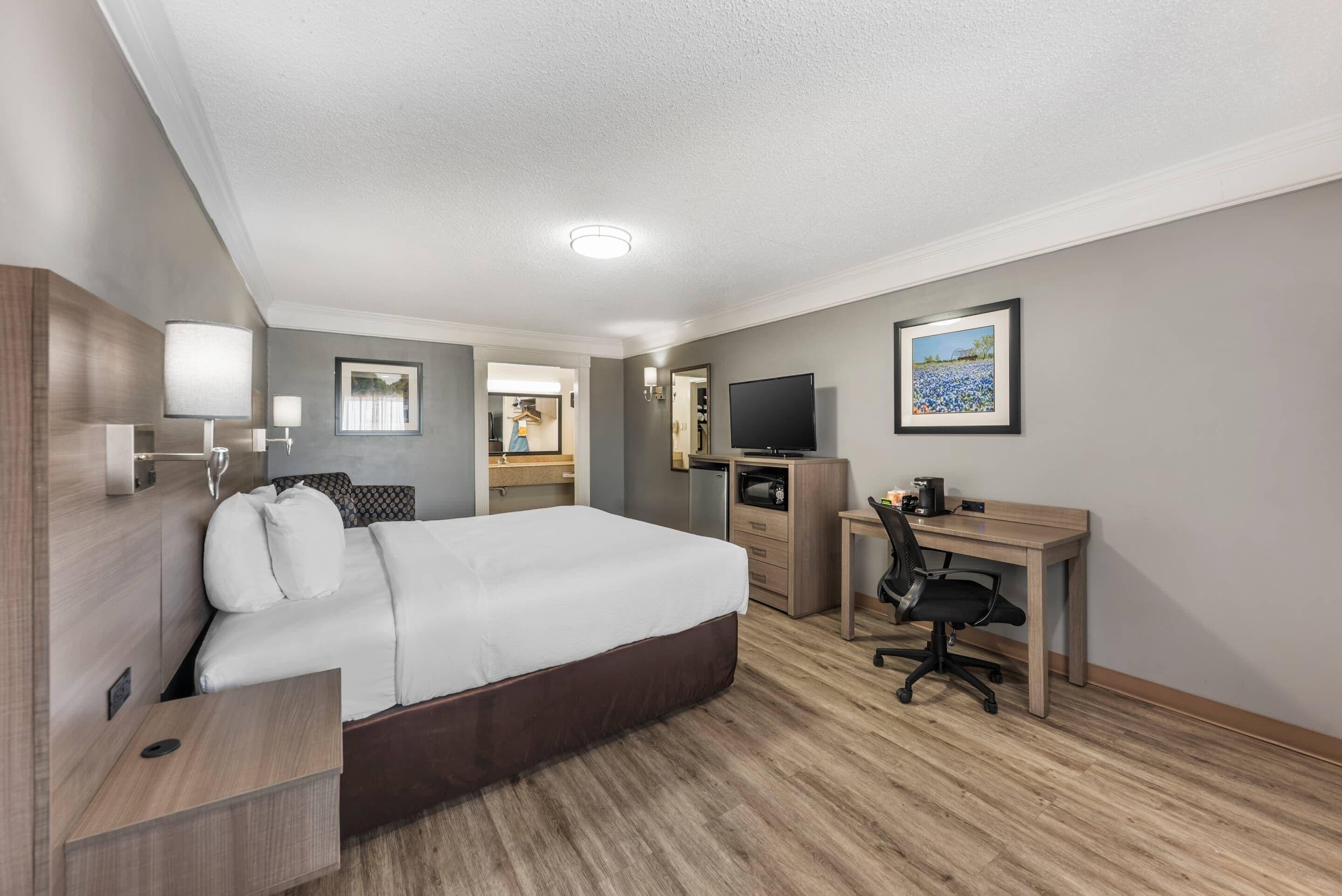 Quarto de hotel com cama, escrivaninha, cadeira, televisão e um quadro na parede.