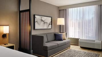 Quarto de hotel moderno com sofá cinza, tapete estampado, obra de arte abstrata emoldurada, luminária de pé e uma janela grande com cortinas.