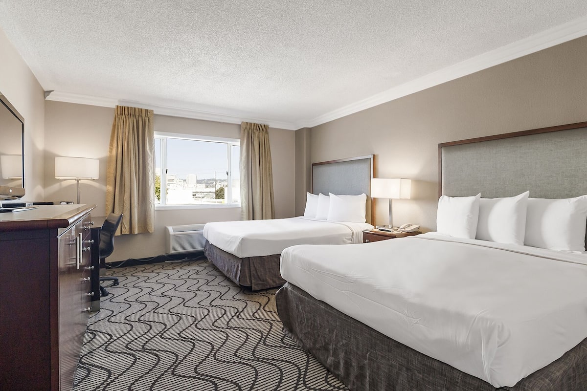 Quarto de hotel com duas camas, uma escrivaninha, uma cadeira, um abajur e vista para a cidade.