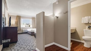 Quarto de hotel com cama, escrivaninha com computador, janela com cortinas, banheiro com vaso sanitário e pia, e chuveiro.