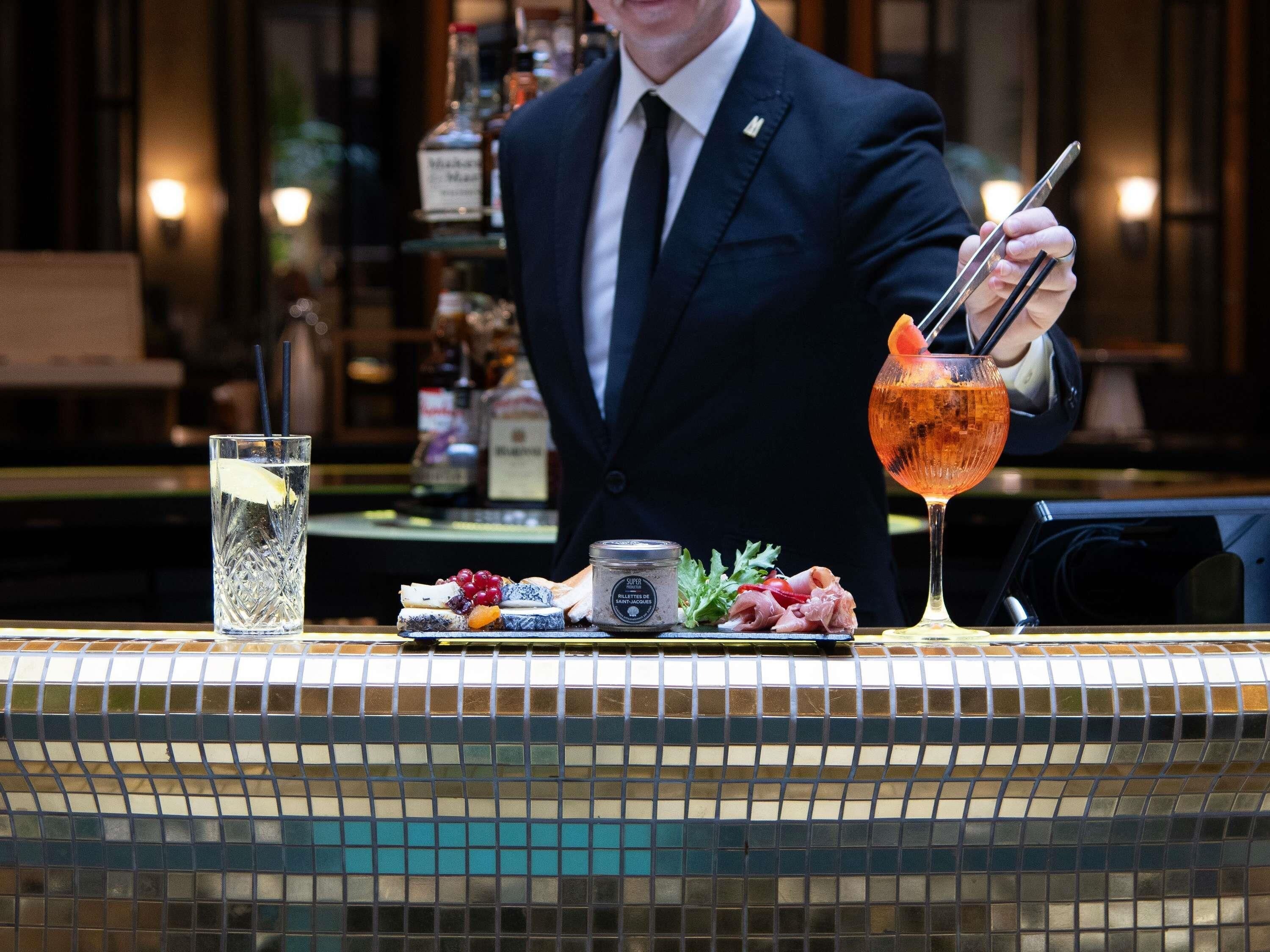 Um bar com um bartender, um copo de água, um cocktail e uma seleção de queijos e enchidos no balcão.