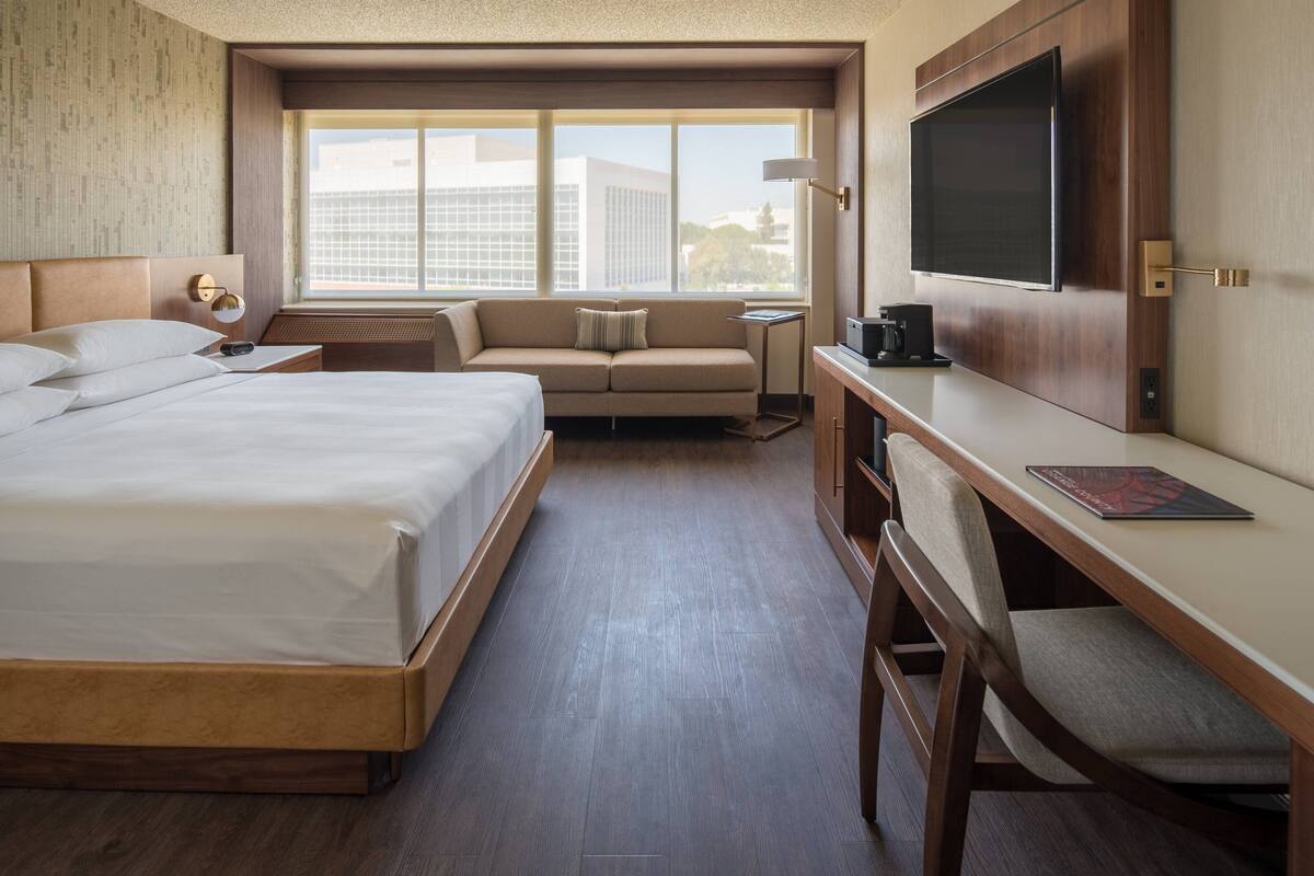 Quarto de hotel com uma cama grande, um sofá, uma escrivaninha com cadeira, uma televisão fixada na parede e uma janela com vista para edifícios.