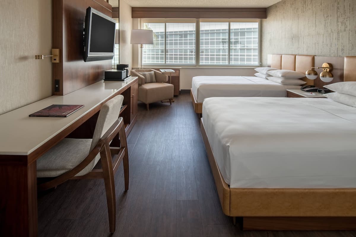 Quarto de hotel com escrivaninha, duas camas, televisão e janela com persianas.