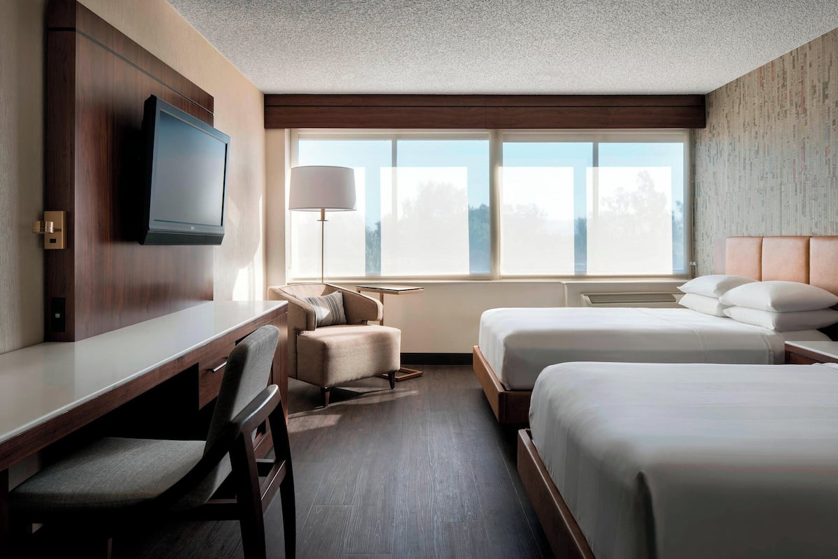 Quarto de hotel com escrivaninha, duas camas, televisão e janela com vista.