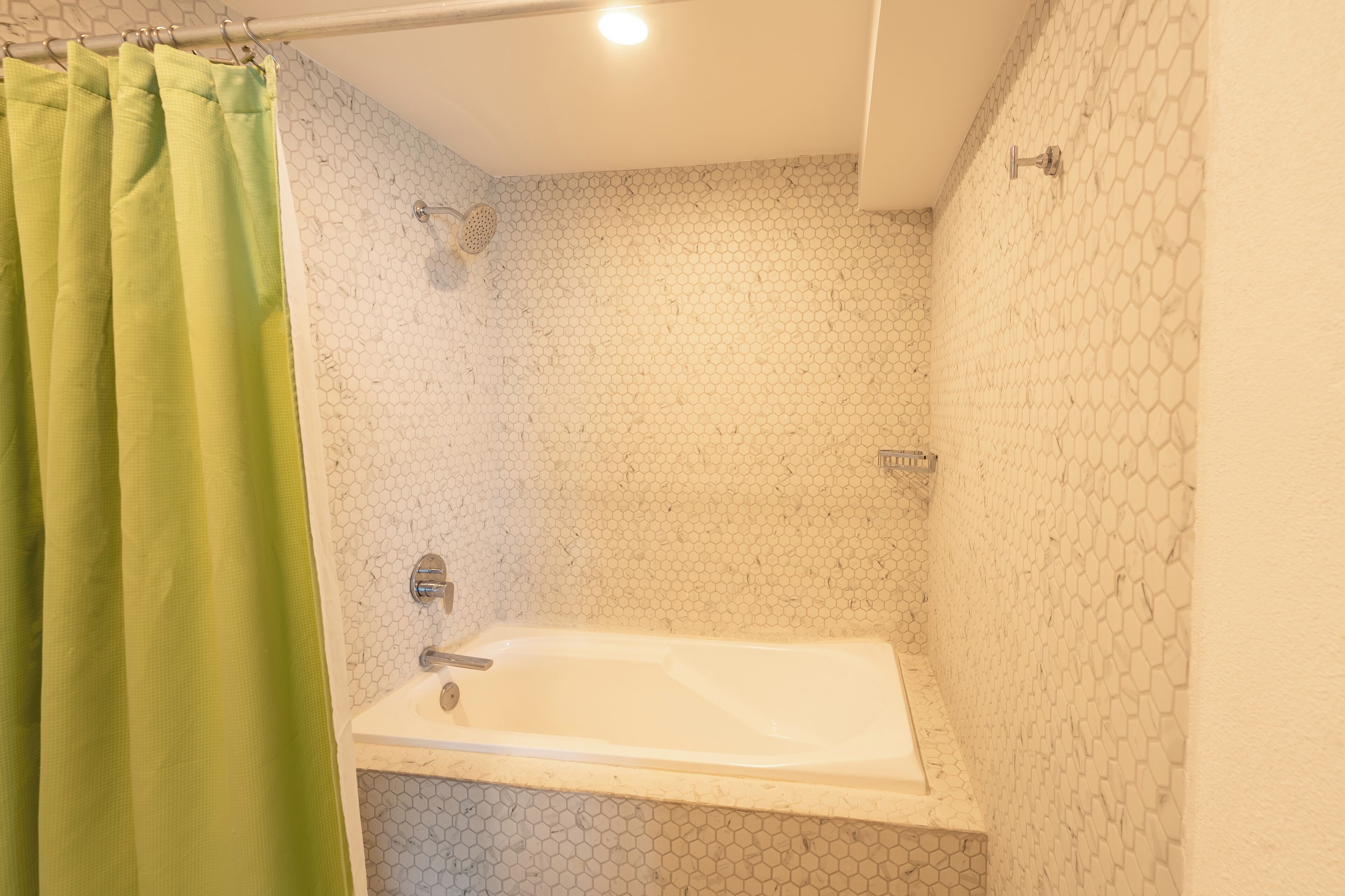 Um banheiro com parede de azulejos hexagonais, uma banheira branca e um cortina de chuveiro verde.