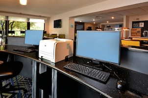 Espaço de escritório moderno com várias estações de trabalho, cada uma equipada com computador, teclado e monitor. Há uma impressora no balcão, e o espaço possui grandes janelas que permitem entrada de luz natural.