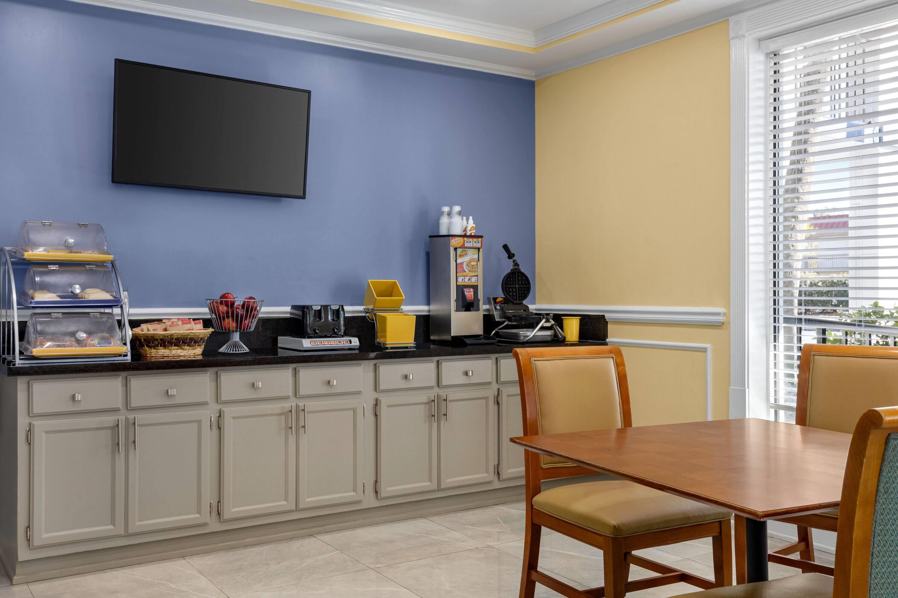 Quarto de hotel com cozinha compacta, área de refeições com mesa e cadeiras, e uma grande janela com persianas.