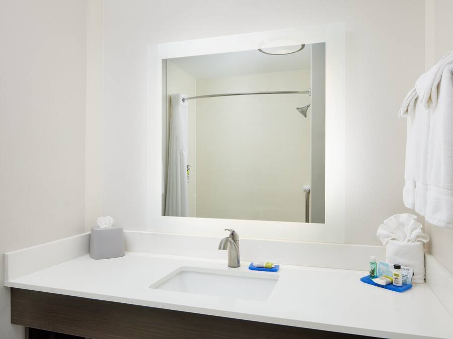 Casa de banho de hotel com lavatório branco, espelho, porta-toalhas e artigos de higiene no balcão.